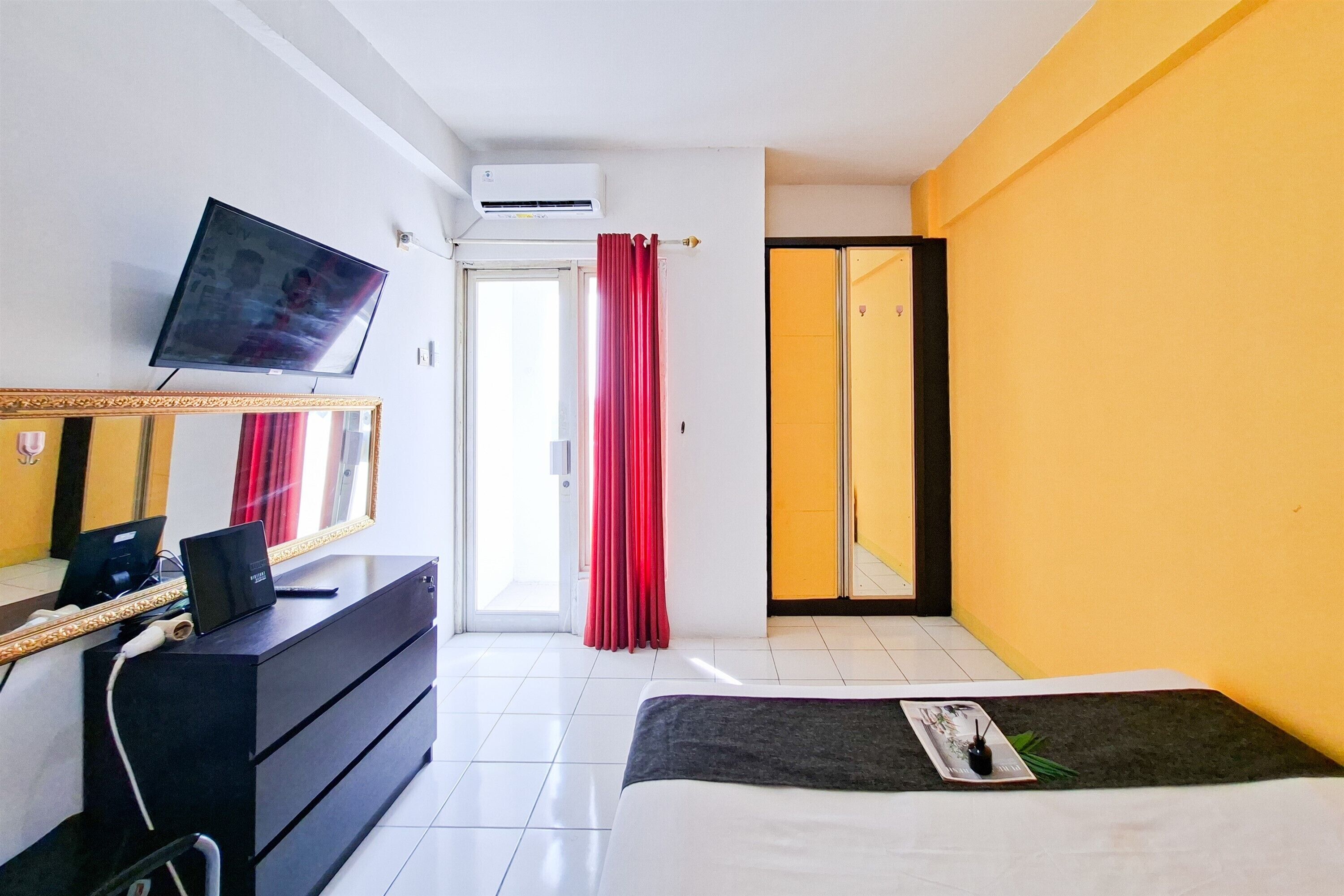 Quarto de hotel com cama, escrivaninha com computador, televisão, espelho e porta de correr para uma varanda.