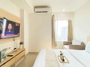 Quarto de hotel com cama, televisão na parede, janela com cortinas e pequena mesa com bandeja.