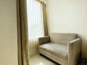 Quarto de canto com sofá bege, janela com cortinas leves e parede branca sem detalhes.
