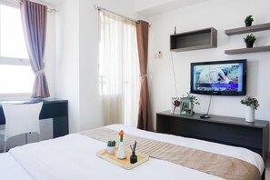 Quarto de hotel com cama, escrivaninha, televisão e uma pequena planta na parede.
