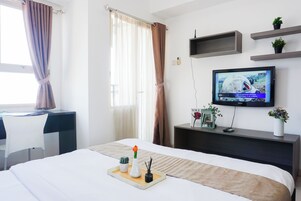 Quarto de hotel com cama, escrivaninha, televisão e uma pequena planta na parede.