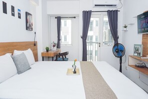 Quarto de hotel com cama, escrivaninha, cadeira, janela com cortinas, ventoinha e televisão na parede.