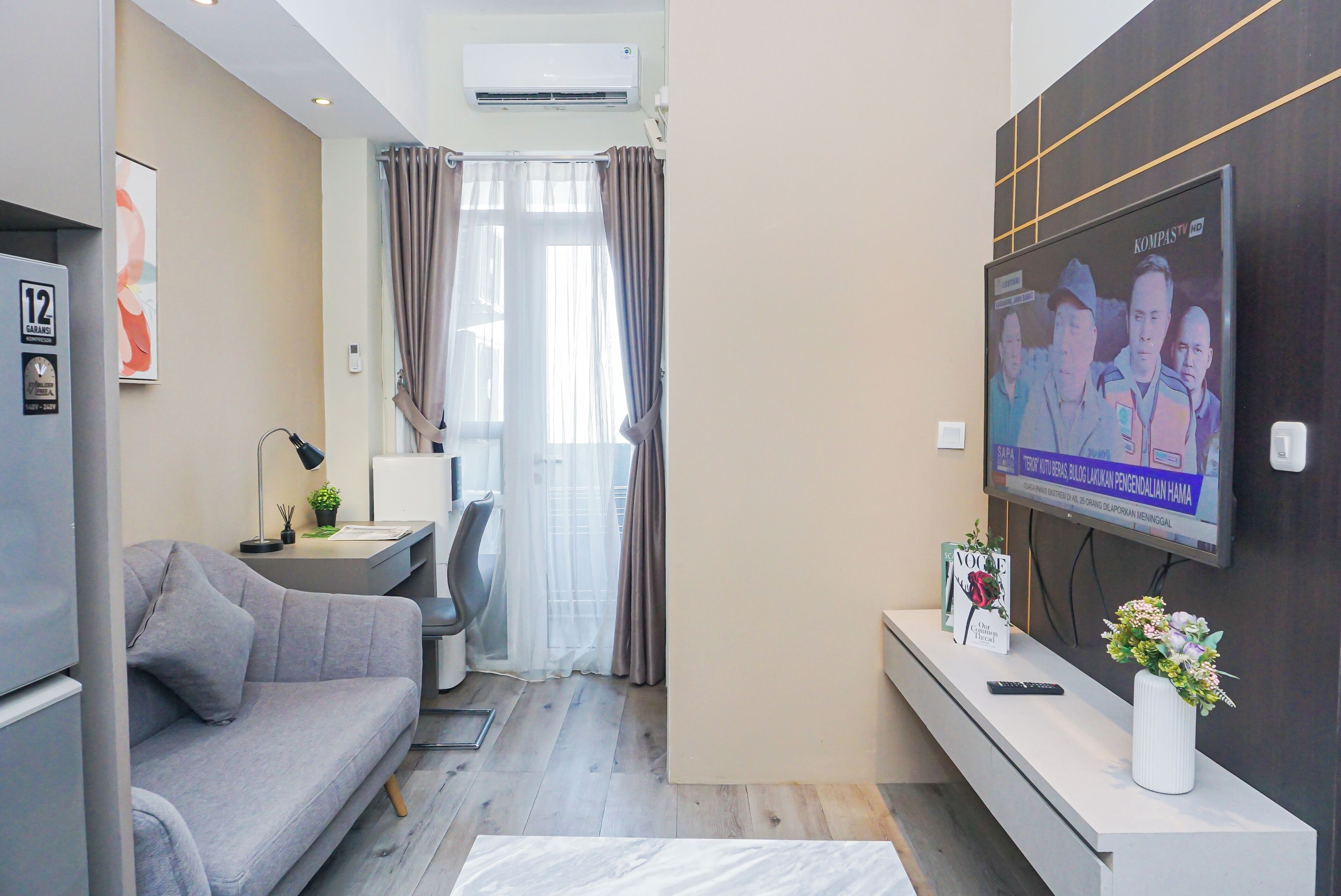 Uma sala de estar moderna com um sofá cinza, uma mesinha com abajur, uma TV fixada na parede e um vaso de planta em uma mesinha lateral.