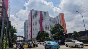 Um edifício de vários andares com uma faixa rosa marcante, rodeado por carros estacionados e vegetação.