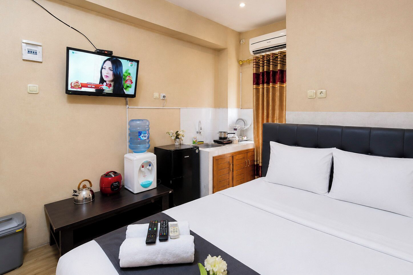 Quarto de hotel com cama, TV fixada na parede, dispensador de água, chaleira, lixeira e uma pequena mesa com controle remoto e toalha.