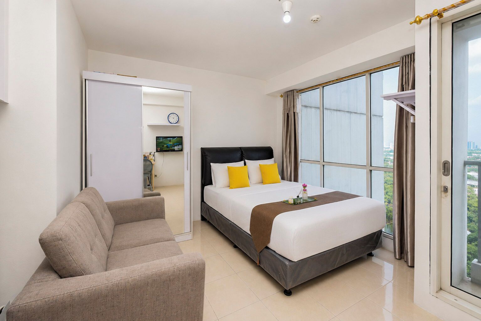 Quarto de hotel com cama, sofá, televisão e vista da cidade através de grandes janelas.