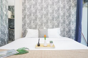 Uma cama bem arrumada com roupa de cama branca, um criado-mudo com um vaso e uma pequena planta, e uma revista no chão.