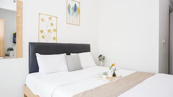 Uma cama bem arrumada com roupa de cama branca e uma cabeceira escura, uma mesa de cabeceira com um vaso e um livro, e um espelho fixado na parede.