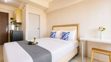 Quarto de hotel com uma cama, uma pequena mesa, uma cadeira, um frigorífico e uma porta.