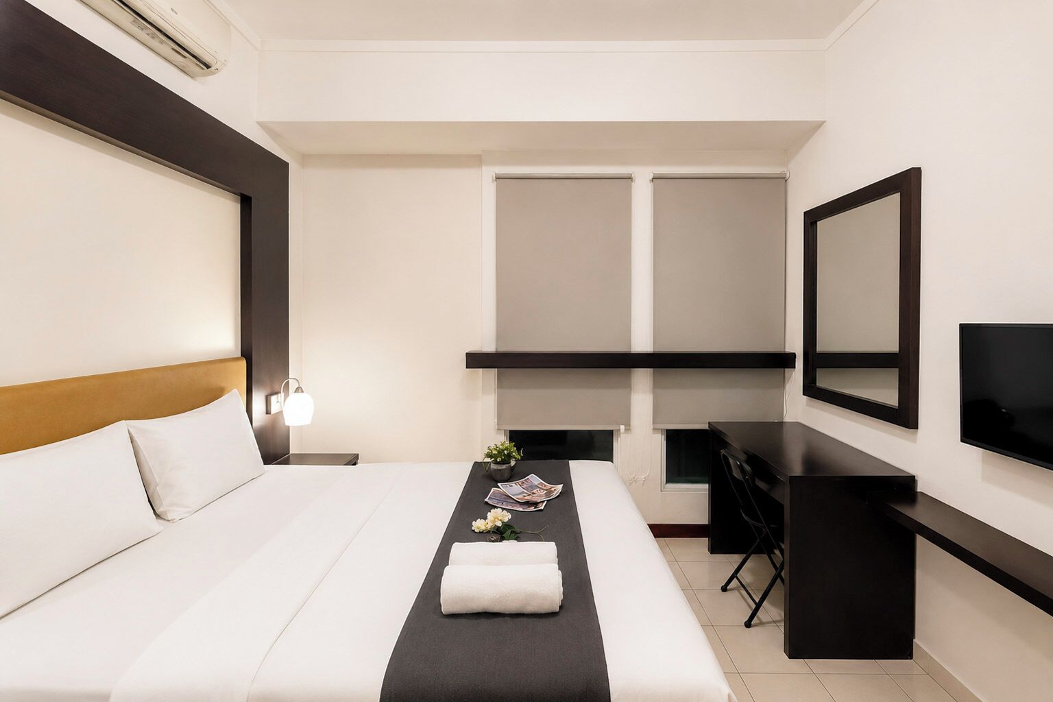 Quarto de hotel com cama, mesa de cabeceira, abajur, escrivaninha, cadeira e televisão.