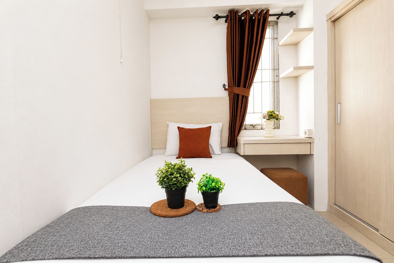 Uma cama de solteiro com cabeceira, duas plantas em vasos, um cobertor cinza, cortina marrom e um guarda-roupa de madeira.