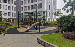 Um parquinho com escorrega e estruturas de escalada, cercado por um jardim ajardinado e um edifício ao fundo.