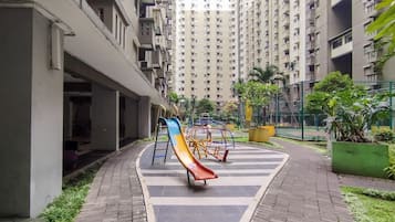 Um parquinho com escorregadores e balanços, cercado por uma calçada e áreas verdes, em uma área residencial com edifícios altos.