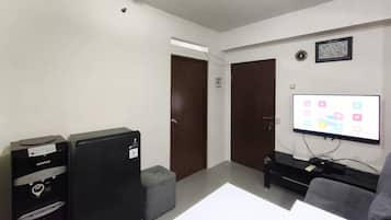 Quarto com televisão de tela plana na parede, móvel de entretenimento preto, um banquinho cinza e uma porta que dá para outro cômodo.