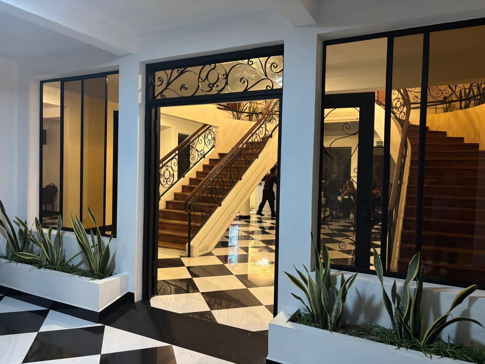 Um hall de hotel com piso xadrez, escada com grades ornamentadas e plantas em vasos.