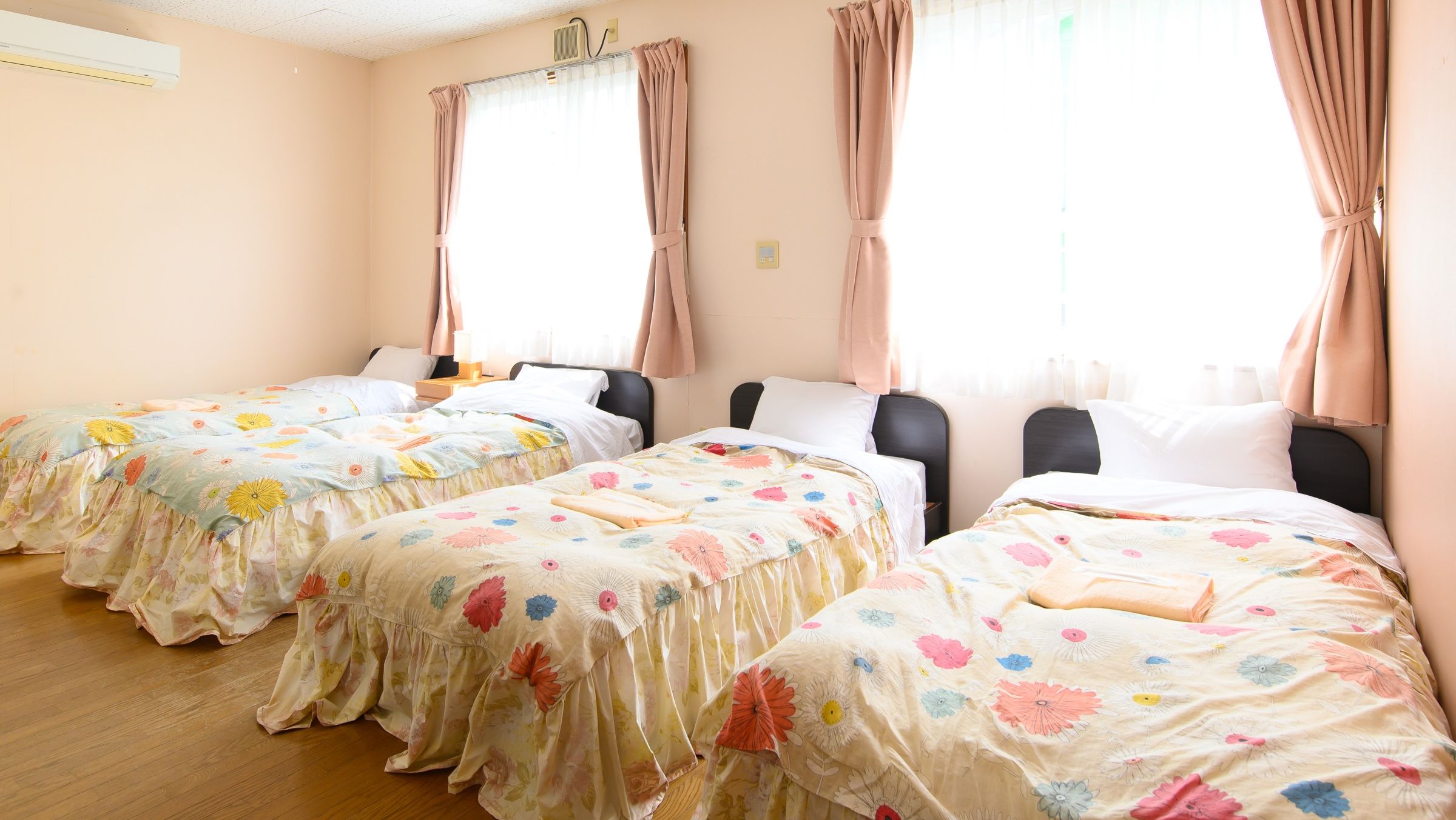 Quarto de hotel com três camas, cada uma com roupa de cama floral, uma mesa de cabeceira com abajur e uma janela com cortinas.
