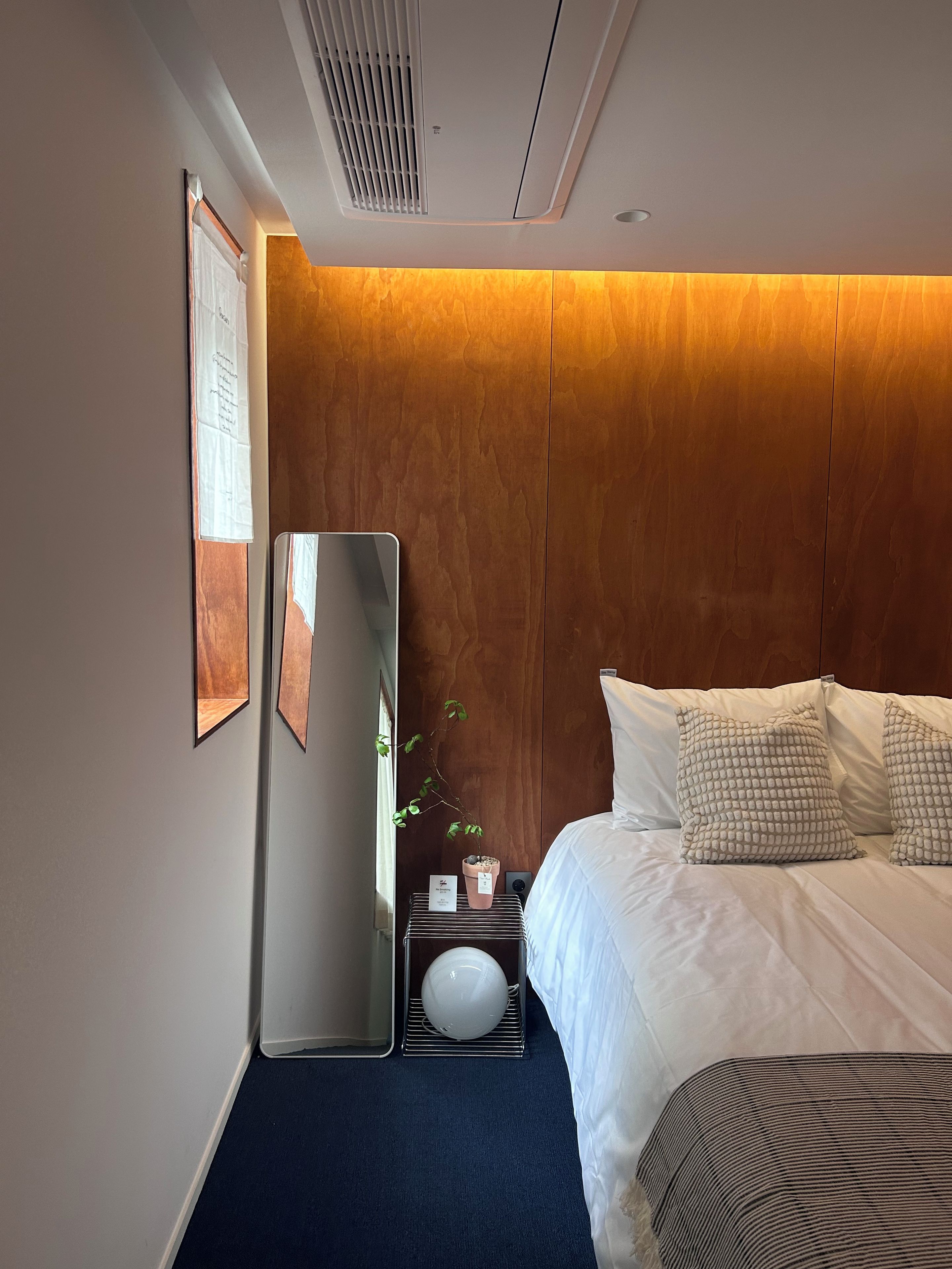 Quarto de hotel moderno com cama, mesa de cabeceira, espelho e uma pequena planta em vaso.