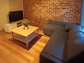 Uma sala de estar com parede de tijolos, televisão de tela plana, mesa de centro de madeira e sofá cinza.