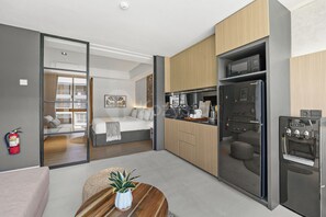 Quarto de hotel moderno com cama, mesa de jantar com uma planta, área de cozinha com frigorífico e micro-ondas, e um extintor de incêndio visível.