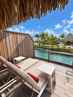Um deck de madeira com espreguiçadeiras e uma pequena mesa, com vista para uma piscina e uma estrutura com telhado de palha ao fundo.