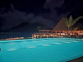 Vista noturna de um resort com piscina, estrutura de telhado de palha e montanha ao fundo.