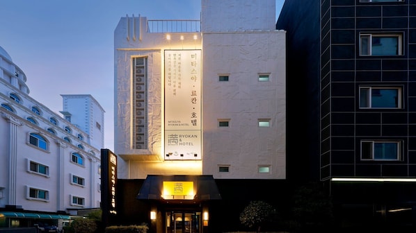 Edifício de hotel com fachada moderna, sinalização indicando 'Ryokan & Hotel' e entrada bem iluminada.