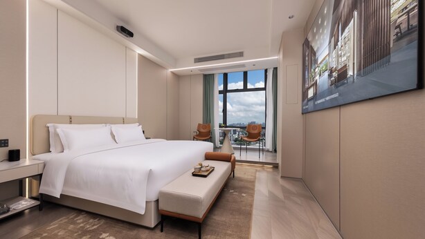 Quarto de hotel moderno com uma cama grande, uma escrivaninha, uma cadeira e vista para a cidade pela janela.