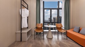 Quarto de hotel moderno com uma janela grande, um sofá, duas cadeiras, uma mesinha e um porta-robes.