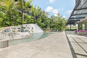Piscina com um muro decorativo, cercada por palmeiras e um edifício moderno com janelas de vidro.