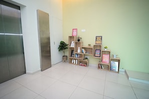 Um corredor com elevador metálico, piso ladrilhado e parede com cartazes motivacionais, prateleiras de madeira com livros e plantas.