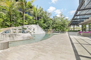 Piscina com um muro decorativo, cercada por palmeiras e um edifício moderno com janelas de vidro.