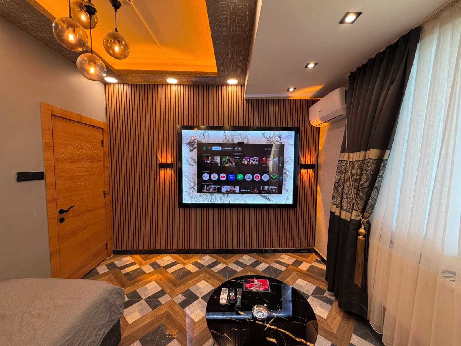 Quarto moderno com televisão de tela plana fixada na parede, porta de madeira, piso estampado e uma pequena mesa com controle remoto.