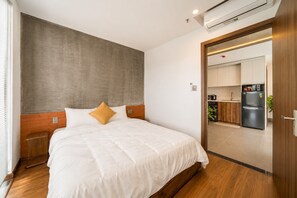 A hotel room with a bed, a wooden headboard, an air conditioning unit, a kitchen area with a refrigerator, and a door leading to another room.