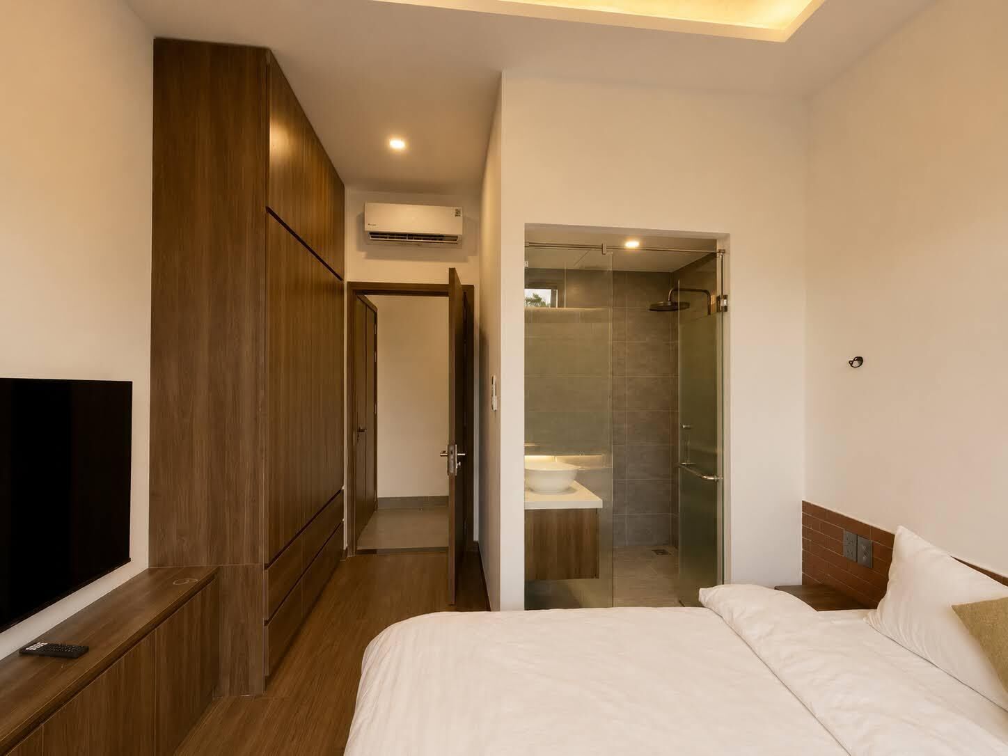 Quarto de hotel moderno com uma cama, uma televisão sobre uma estante de madeira, um guarda-roupa e uma área de chuveiro com paredes de vidro.