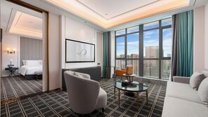 Quarto de hotel com cama, área de estar com mesa, janela ampla com vista para a cidade e uma obra de arte pendurada na parede.
