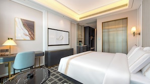 Quarto de hotel com cama, escrivaninha com cadeira, televisão e banheiro visível através de uma porta aberta.