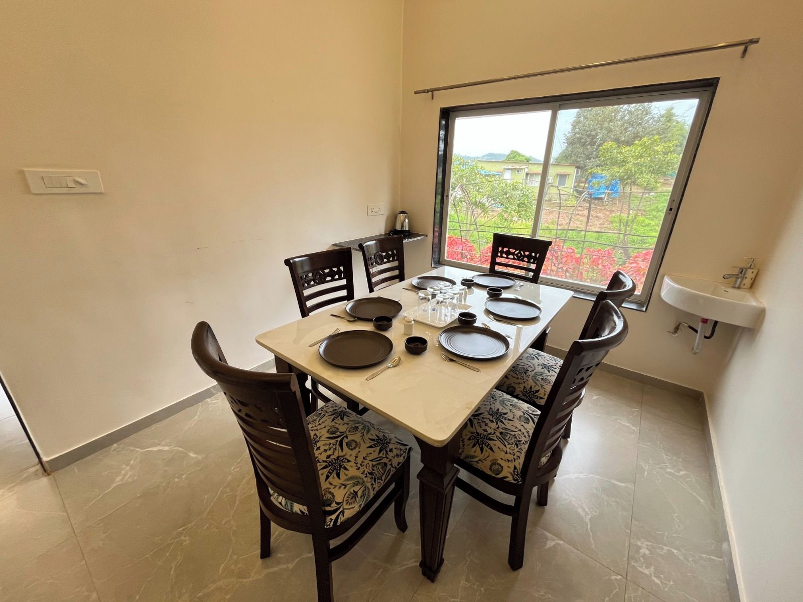 Uma área de refeições com mesa posta para quatro, cadeiras com almofadas estampadas, uma janela com vista para vegetação e uma pia fixa na parede.