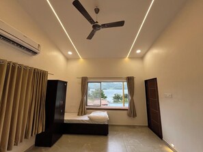 Quarto com cama, ventilador de teto, aparelho de ar condicionado, janela com cortinas e vista para um corpo de água.
