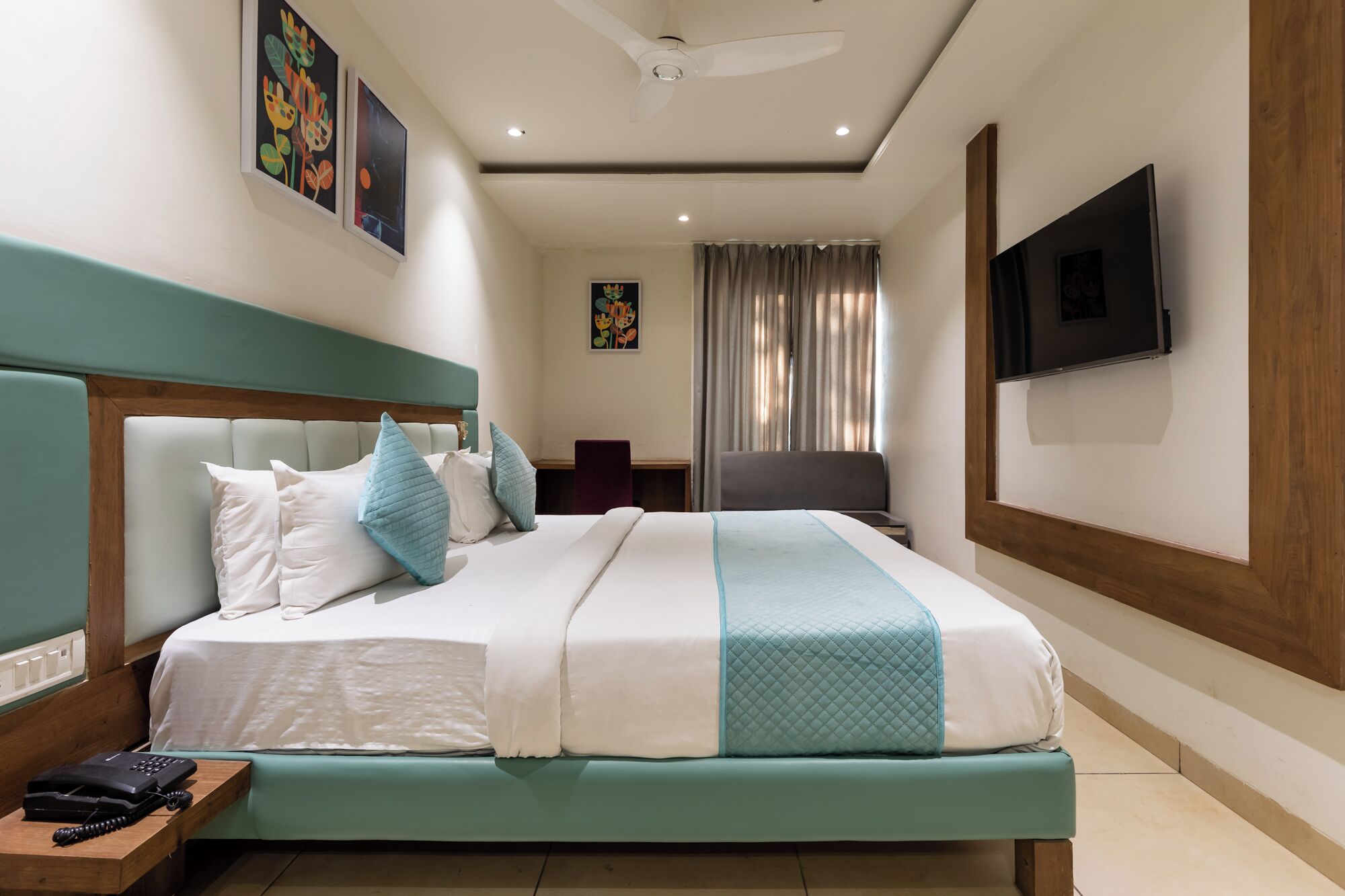 Quarto de hotel com cama, escrivaninha, cadeira, televisão e ventoinha no teto.