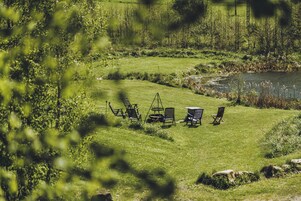 Uma área externa gramada com um lago, cercada por árvores e vegetação. Várias cadeiras e uma mesa estão dispostas para refeições ao ar livre.