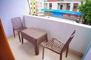 Uma varanda com duas cadeiras de vime e uma mesa, com vista para uma rua com edifícios e uma placa azul do Bank of India.