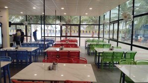 Um restaurante com várias mesas e cadeiras, grandes janelas e vista para palmeiras lá fora.
