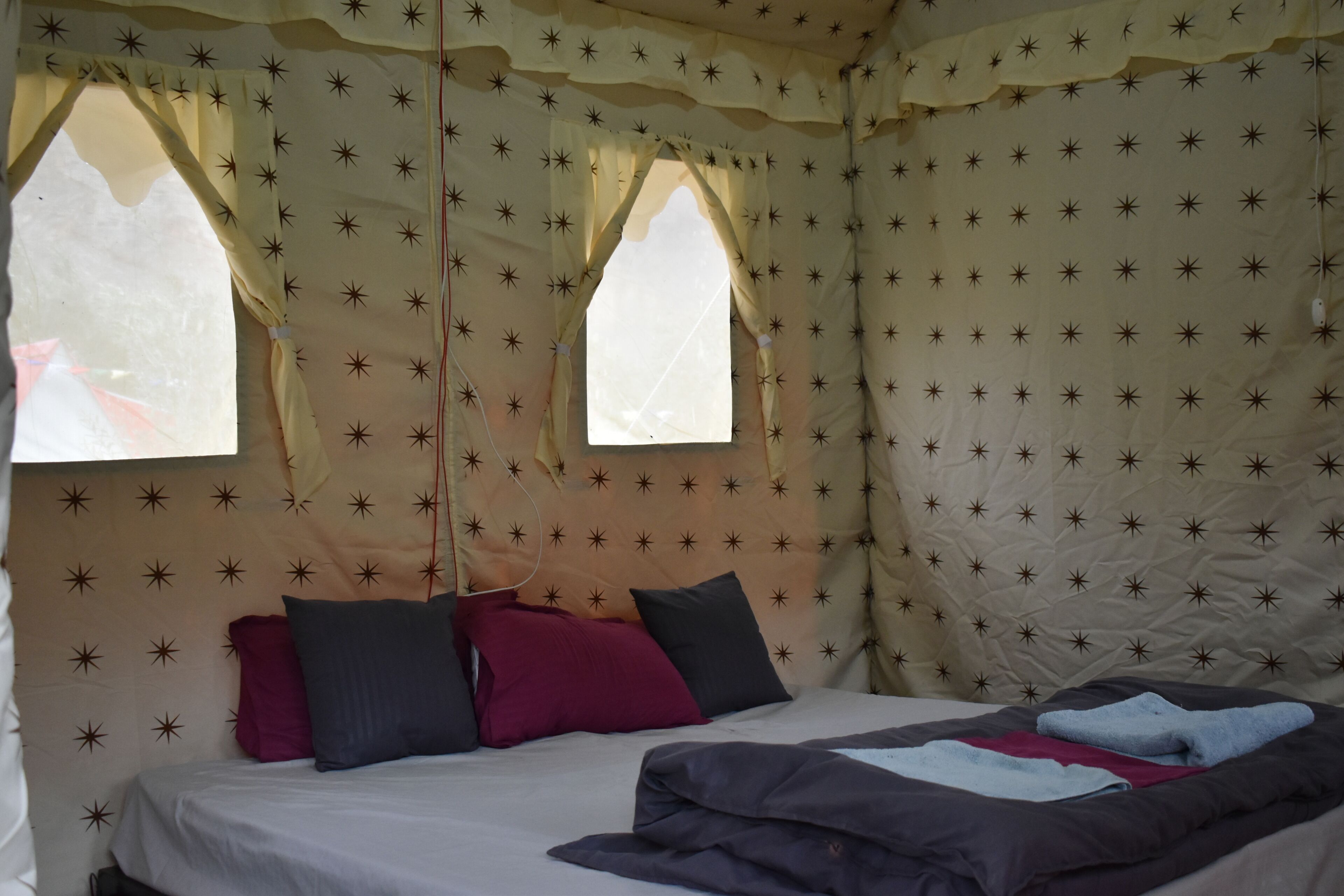 Uma cama com almofadas e um cobertor, uma janela com cortinas e uma parede estampada.