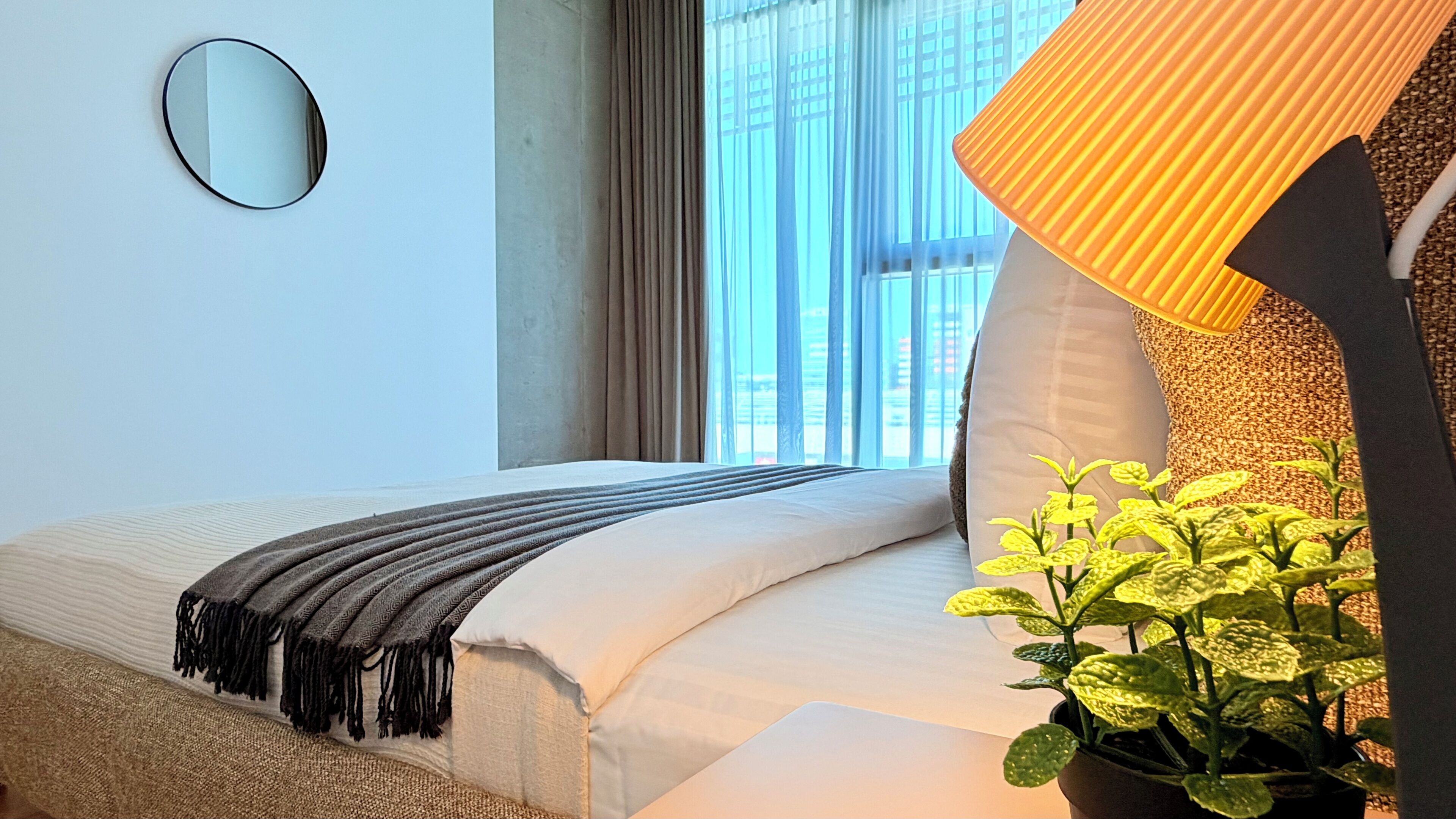 Quarto de hotel com cama, planta em vaso, luminária, espelho e janela com cortinas.