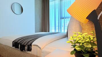 Quarto de hotel com cama, planta em vaso, luminária, espelho e janela com cortinas.