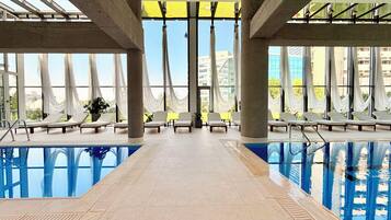 Área da piscina com espreguiçadeiras e vista para edifícios através de grandes janelas.
