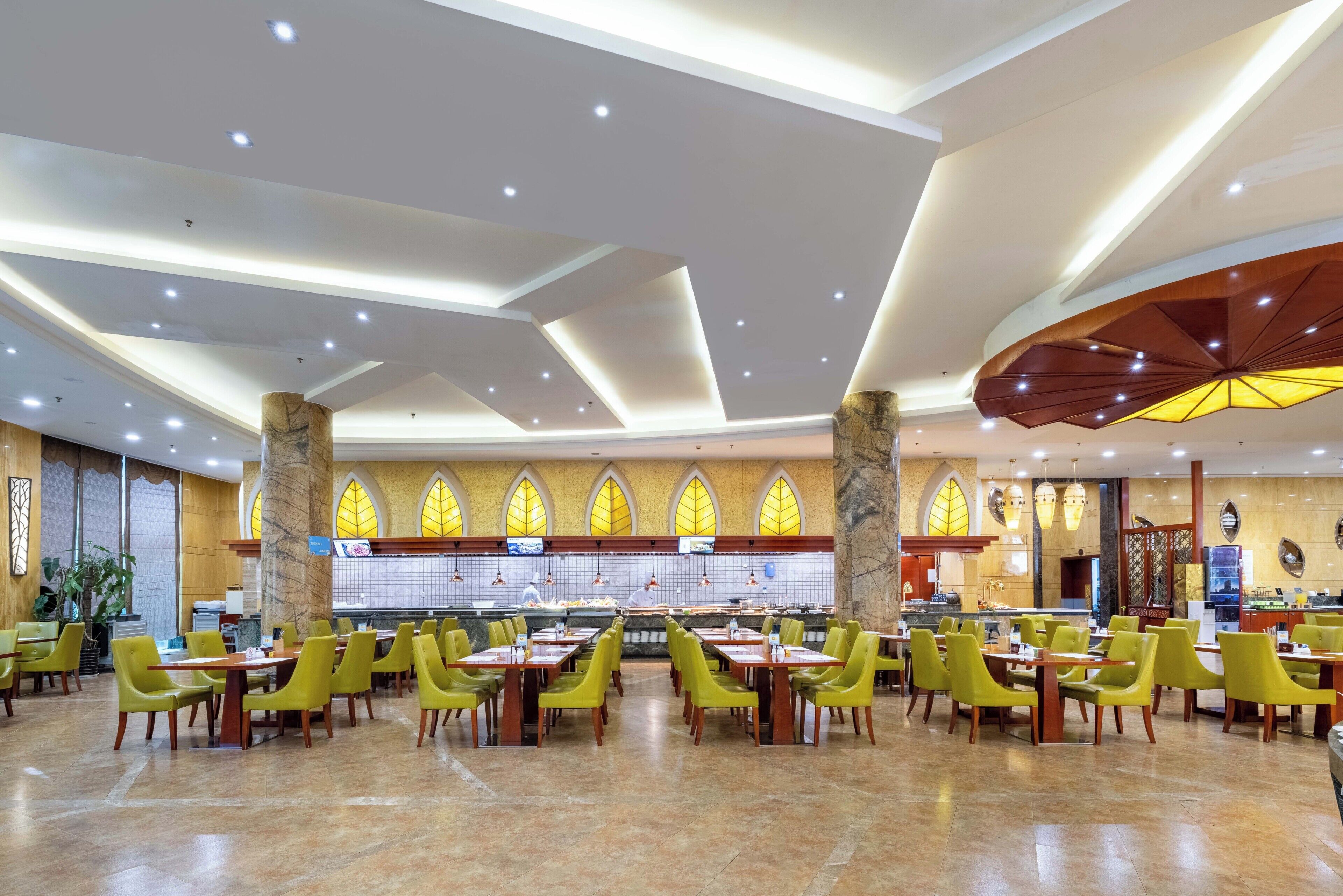 Uma sala de jantar ampla, com várias mesas e cadeiras, uma cozinha ao fundo e um design de teto moderno.