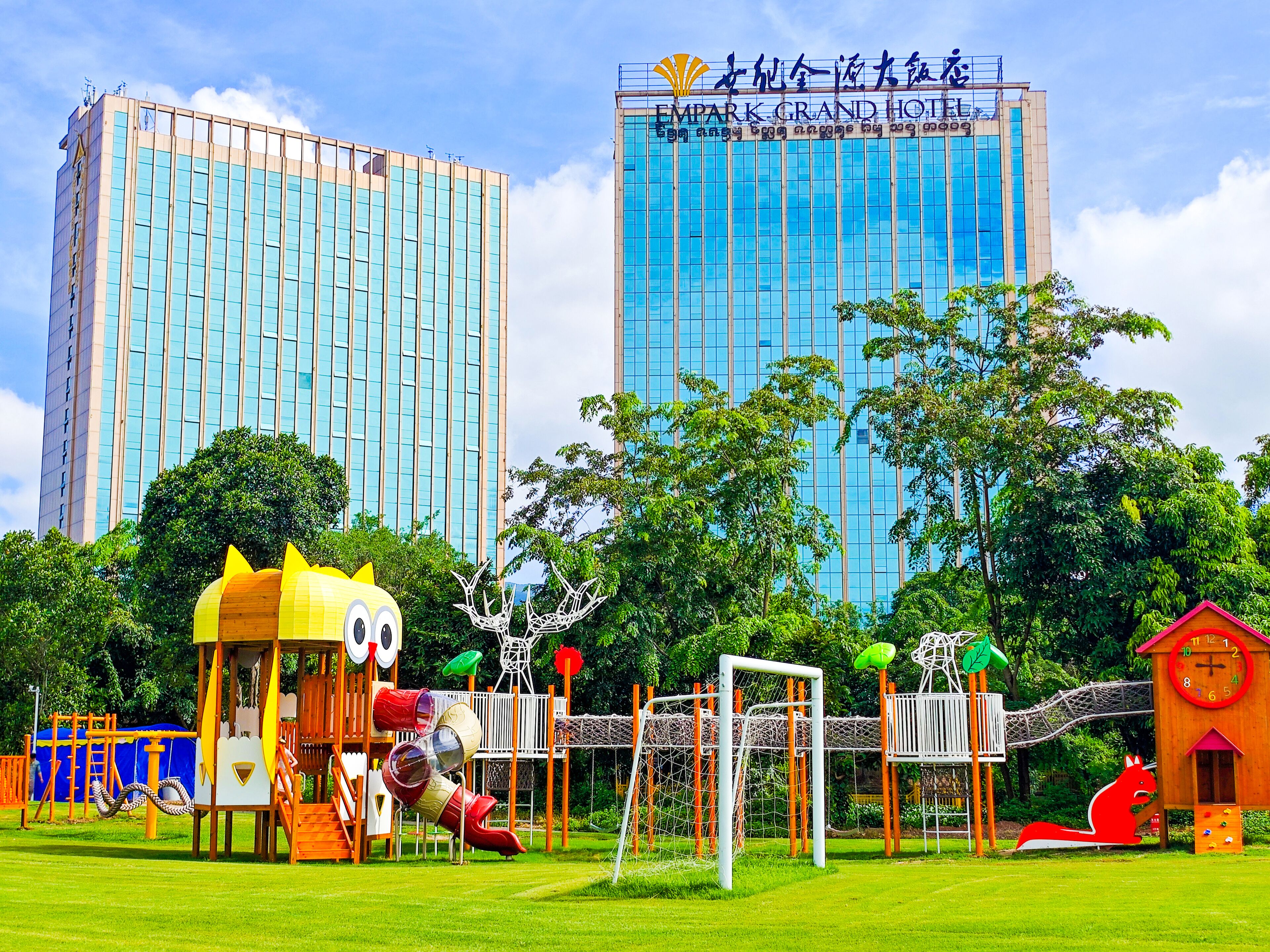 Um parquinho infantil com diversas estruturas de lazer, incluindo um escorrega e uma rede de escalada, em frente a um hotel com fachada azul e placas de sinalização.