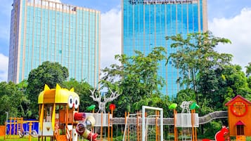 Um parquinho infantil com diversas estruturas de lazer, incluindo um escorrega e uma rede de escalada, em frente a um hotel com fachada azul e placas de sinalização.