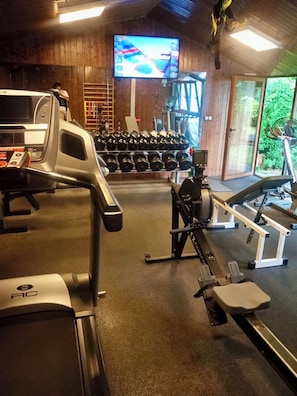 Academia com equipamentos de ginástica, televisão e vista para o verde lá fora.
